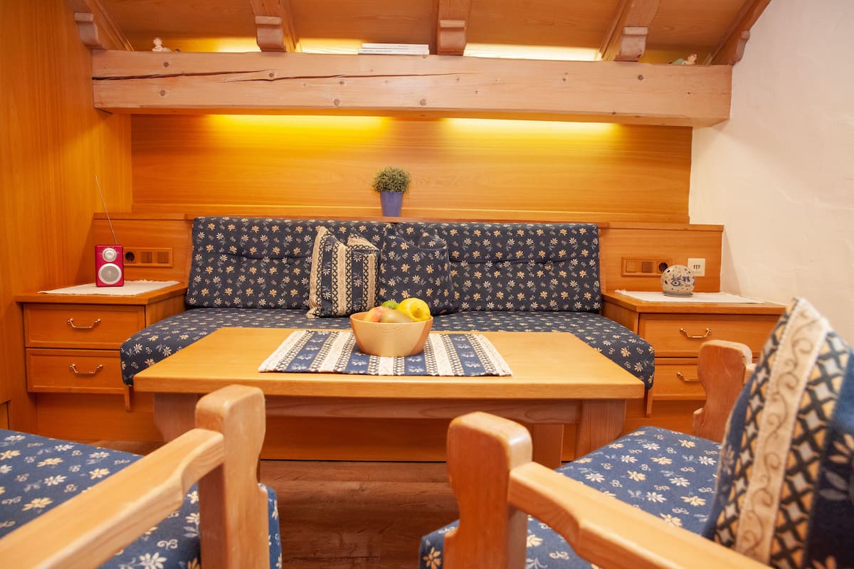 Interior acolhedor de cabana com um banco de madeira, duas cadeiras de madeira, uma mesinha com uma tigela de frutas e um criado-mudo com um rádio.