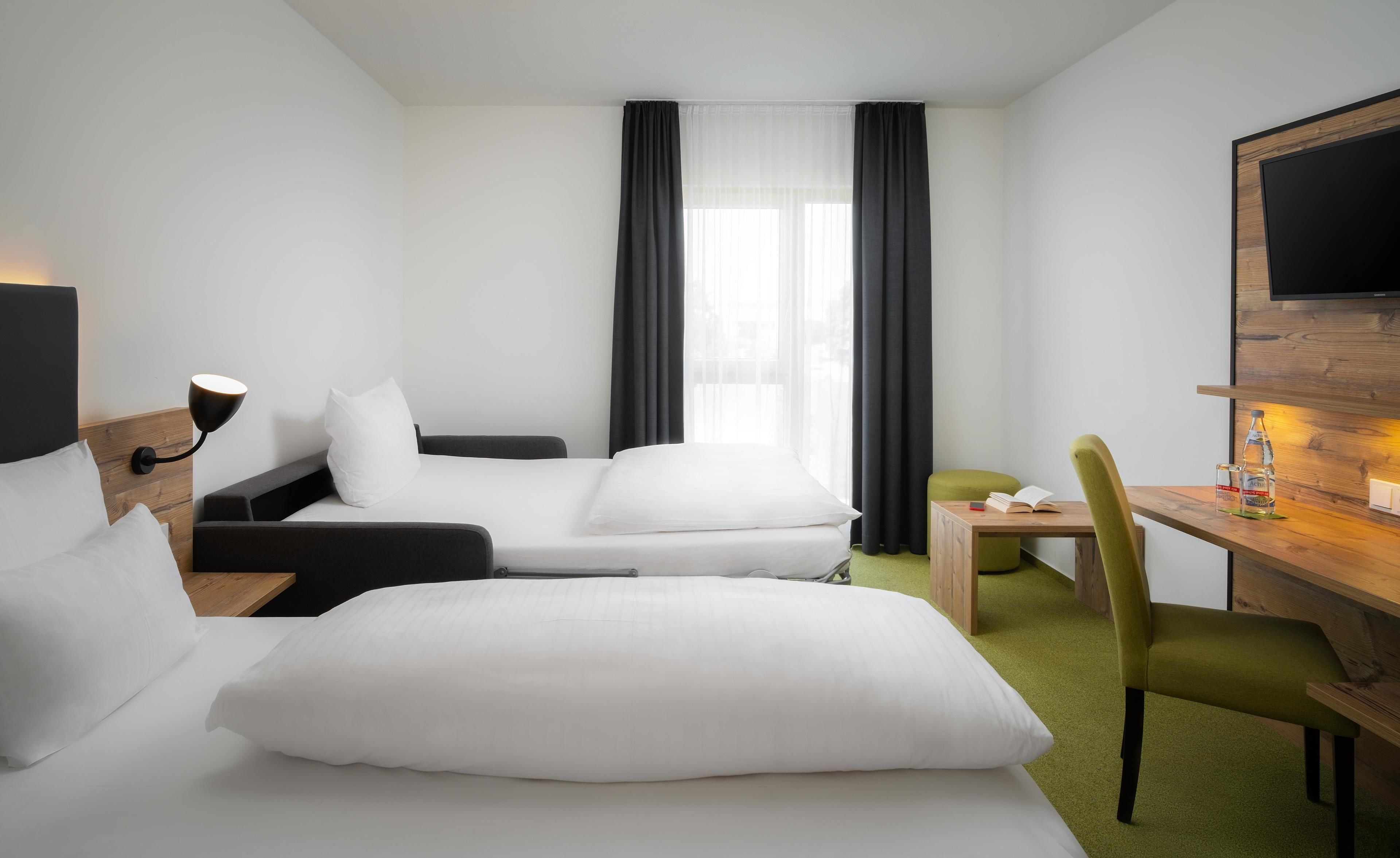 Quarto de hotel com duas camas, uma escrivaninha com cadeira, televisão e janela com cortinas.