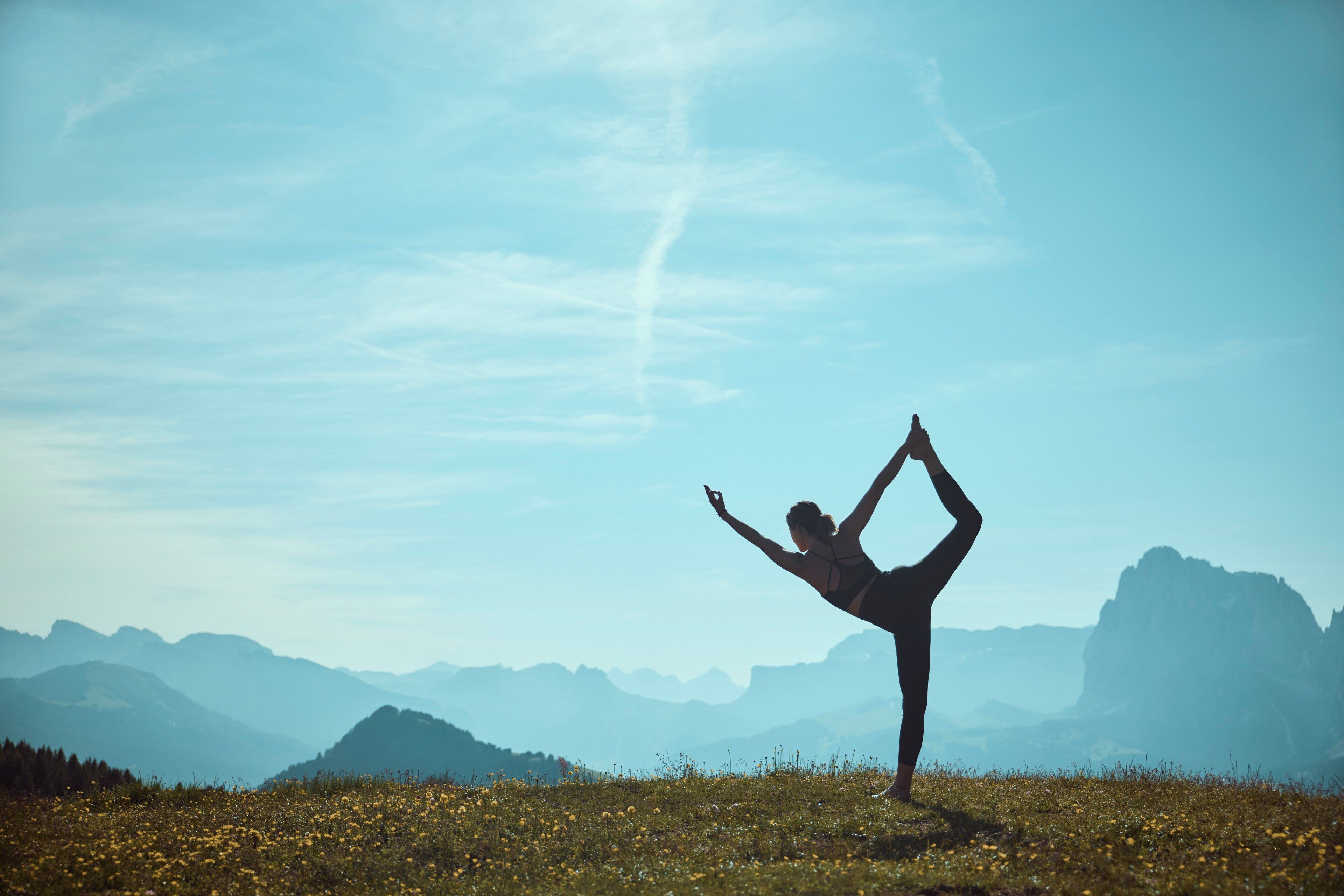 Uma pessoa fazendo uma postura de yoga em um campo gramado, com montanhas ao fundo.