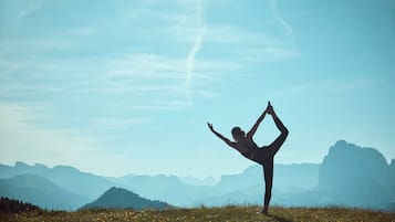 Uma pessoa fazendo uma postura de yoga em um campo gramado, com montanhas ao fundo.