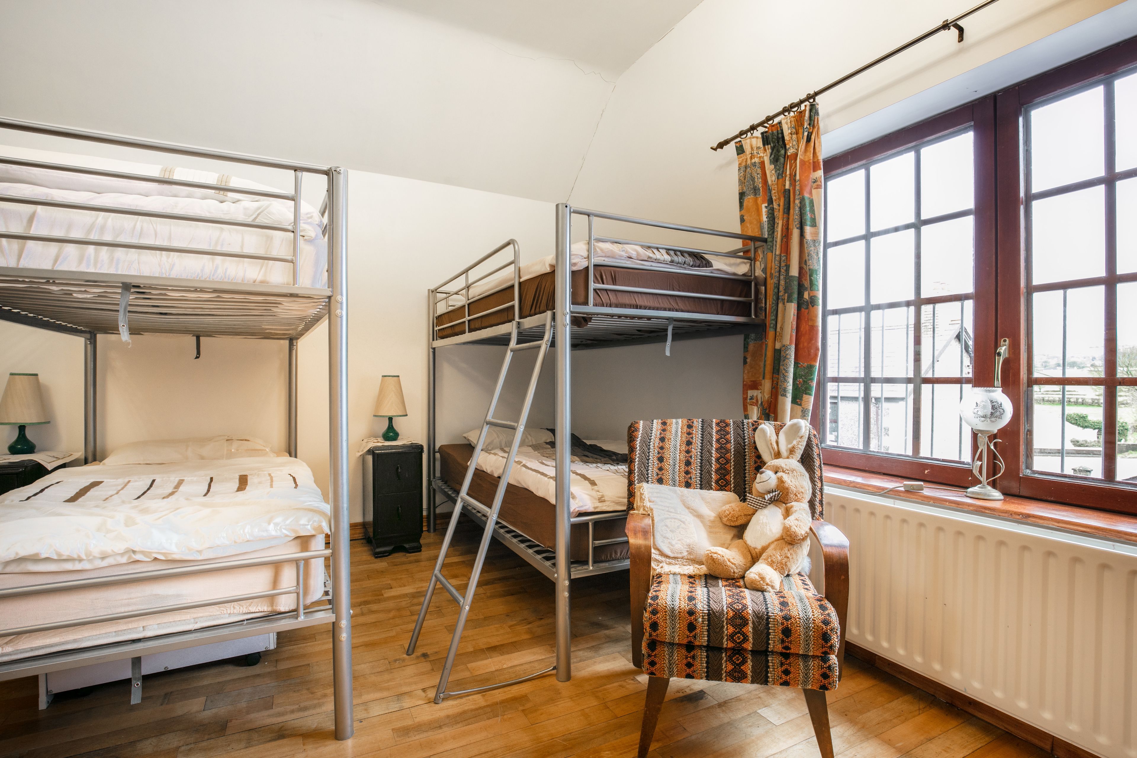 Quarto com beliches, uma cadeira com um brinquedo de pelúcia, um radiador e uma janela com cortinas.
