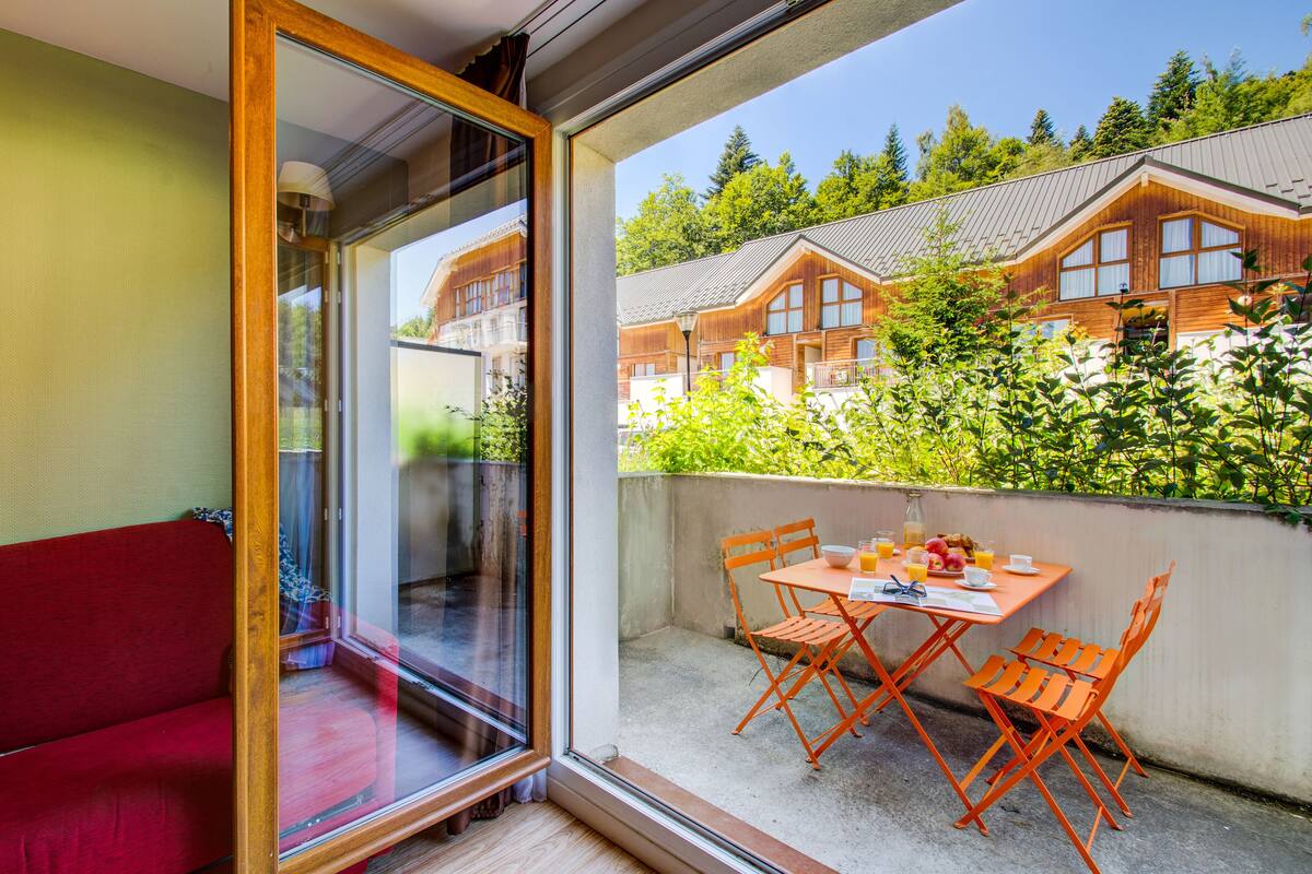 Uma varanda com mesa e cadeiras, um sofá vermelho e vista para edifícios de madeira e vegetação.