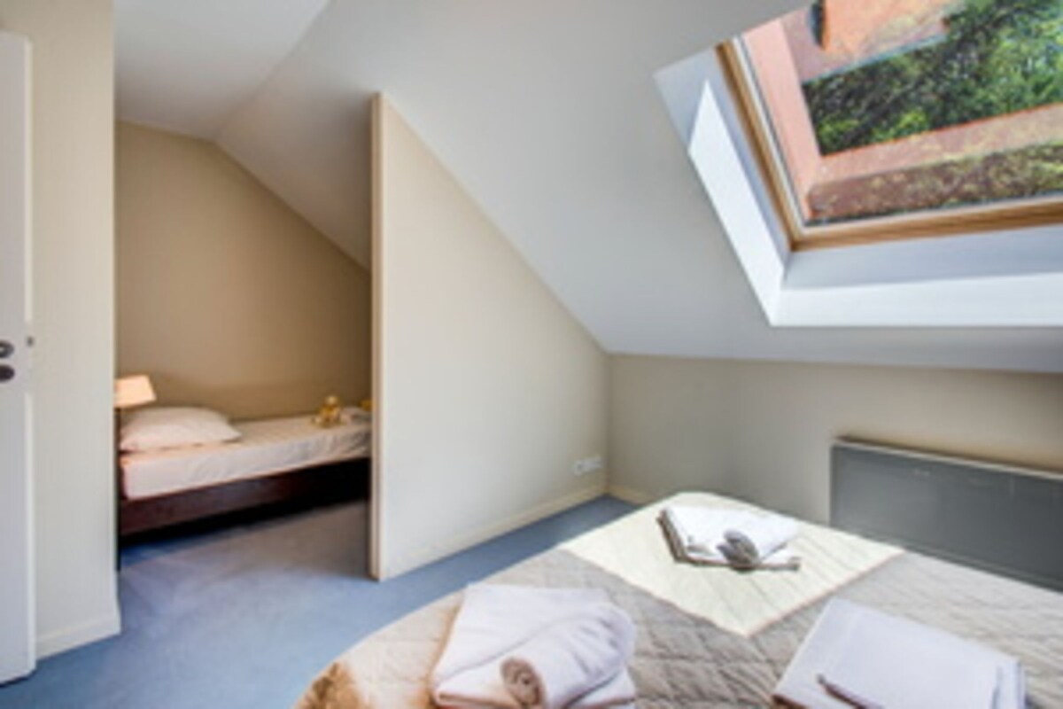 Quarto com teto inclinado, cama, mesa de cabeceira e janela com vista para vegetação.