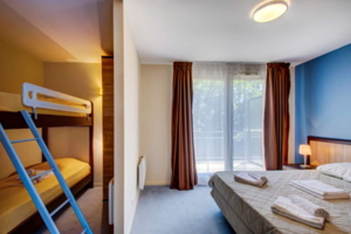 Quarto de hotel com beliches, uma janela grande com cortinas, mesa de cabeceira com luminária e uma cama arrumada.