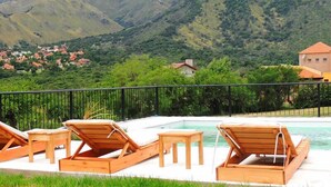 Área de piscina com espreguiçadeiras de madeira e vista para montanhas e uma aldeia.