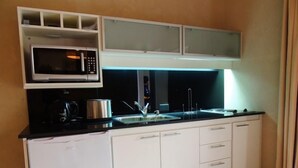 Cozinha compacta com armários brancos, bancada preta, micro-ondas embutido e chaleira de aço inoxidável.
