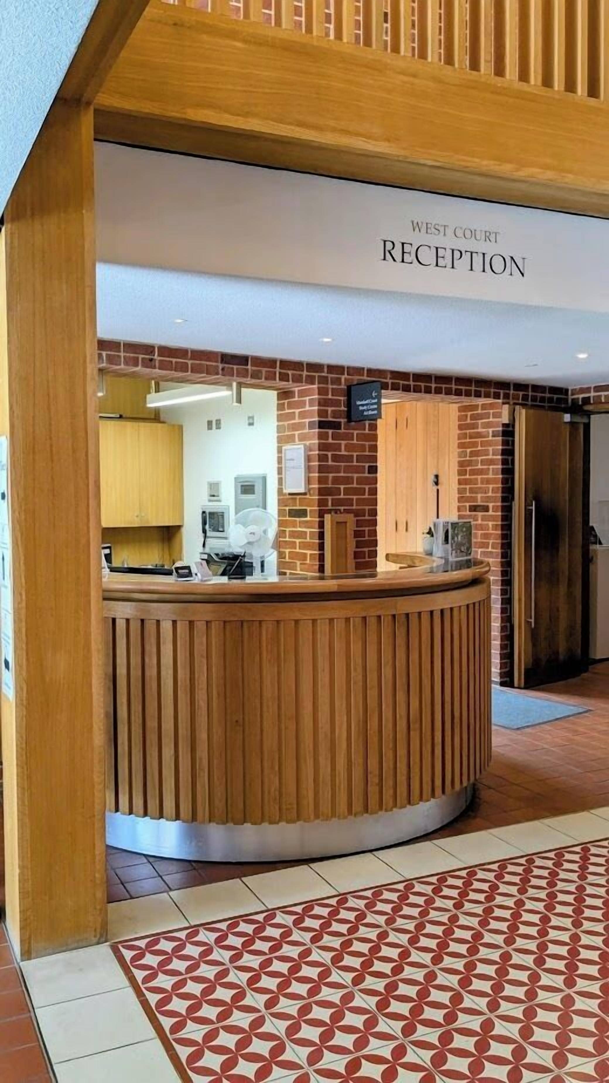 Uma área de recepção com um balcão de madeira, uma placa com a inscrição 'West Court Reception' e piso estampado.