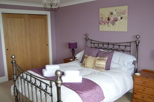 Uma cama bem arrumada com lençóis brancos e detalhes em roxo, uma mesa de cabeceira de madeira com abajur e um aparador de madeira.