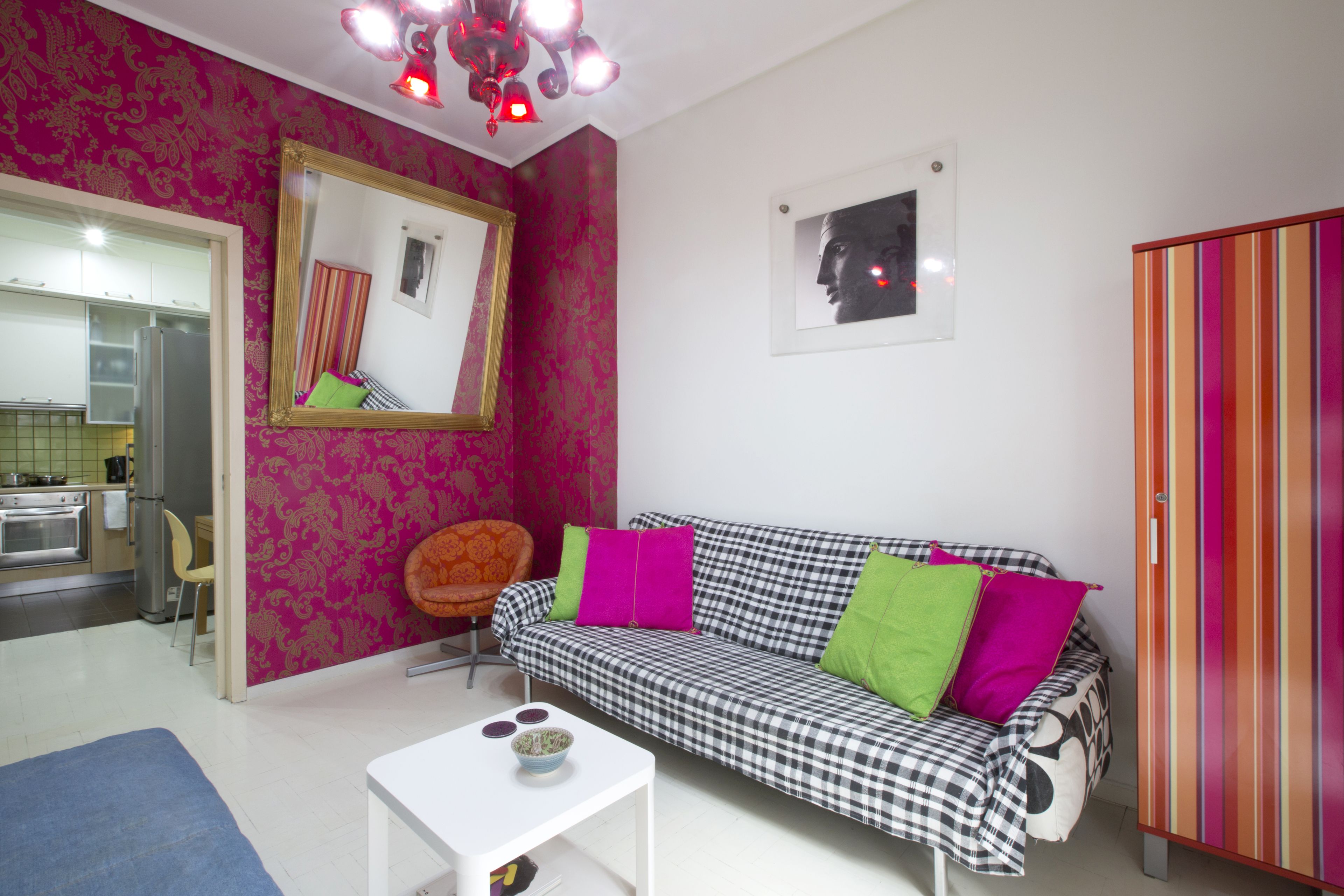 Uma sala de estar com um sofá estampado em xadrez, uma mesa pequena branca, um guarda-roupa colorido com listras, um espelho e uma cozinha ao fundo.