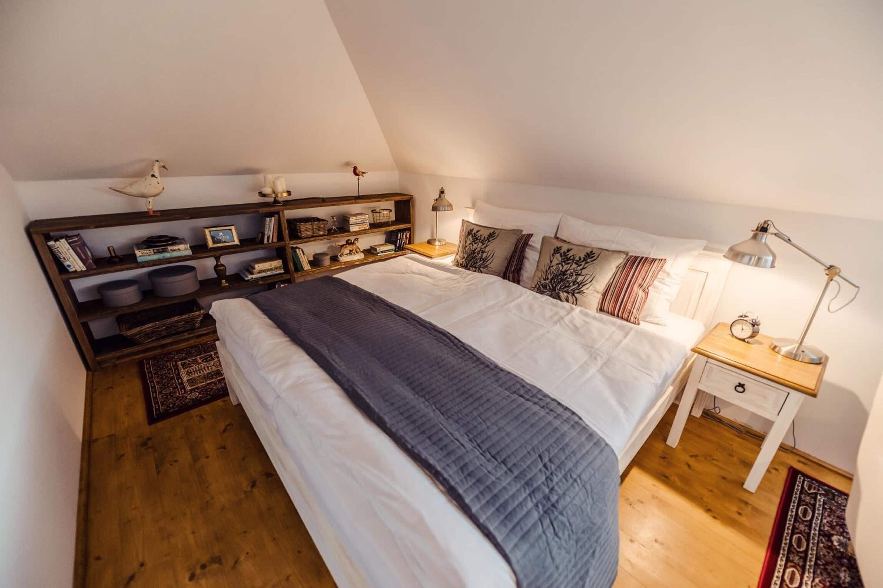 Quarto com cama grande, piso de madeira, estante, mesa de cabeceira com luminária e uma pequena mesa com despertador.