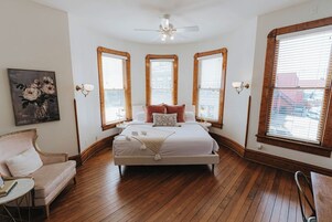 Quarto com uma cama grande, piso de madeira, ventoinha no teto e três janelas com molduras de madeira.
