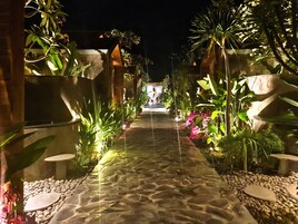 Um caminho noturno com lajes de pedra, rodeado por diversas plantas e uma fonte de luz central.