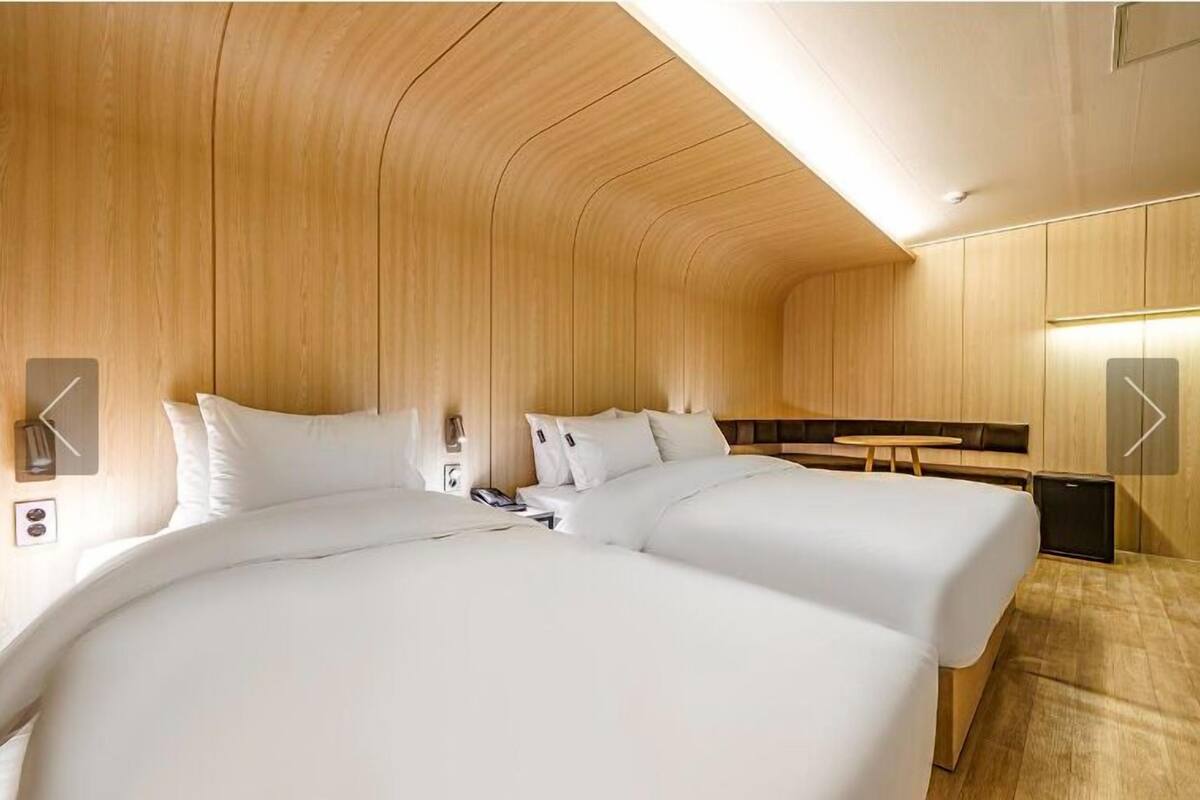 Quarto de hotel moderno com teto curvo de madeira, uma cama grande, mesas de cabeceira e um pequeno espaço de estar.