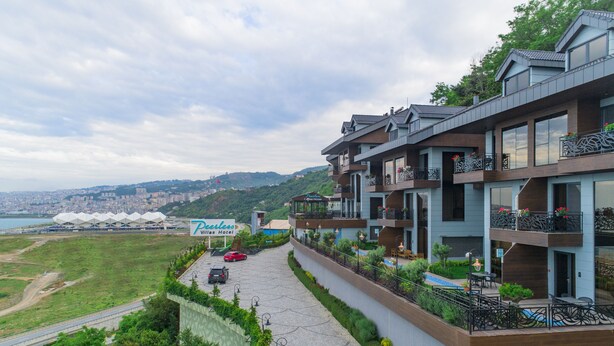 Hotel com várias varandas, um caminho pavimentado e vista para a paisagem urbana e colinas ao fundo.
