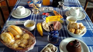 Mesa de café da manhã com diversos alimentos e bebidas, incluindo pão, doces e uma sobremesa de chocolate.