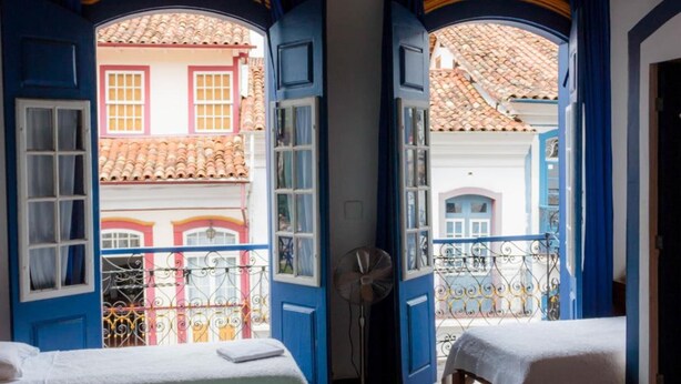 Quarto com duas camas, um ventilador e vista para edifícios através de portas abertas.