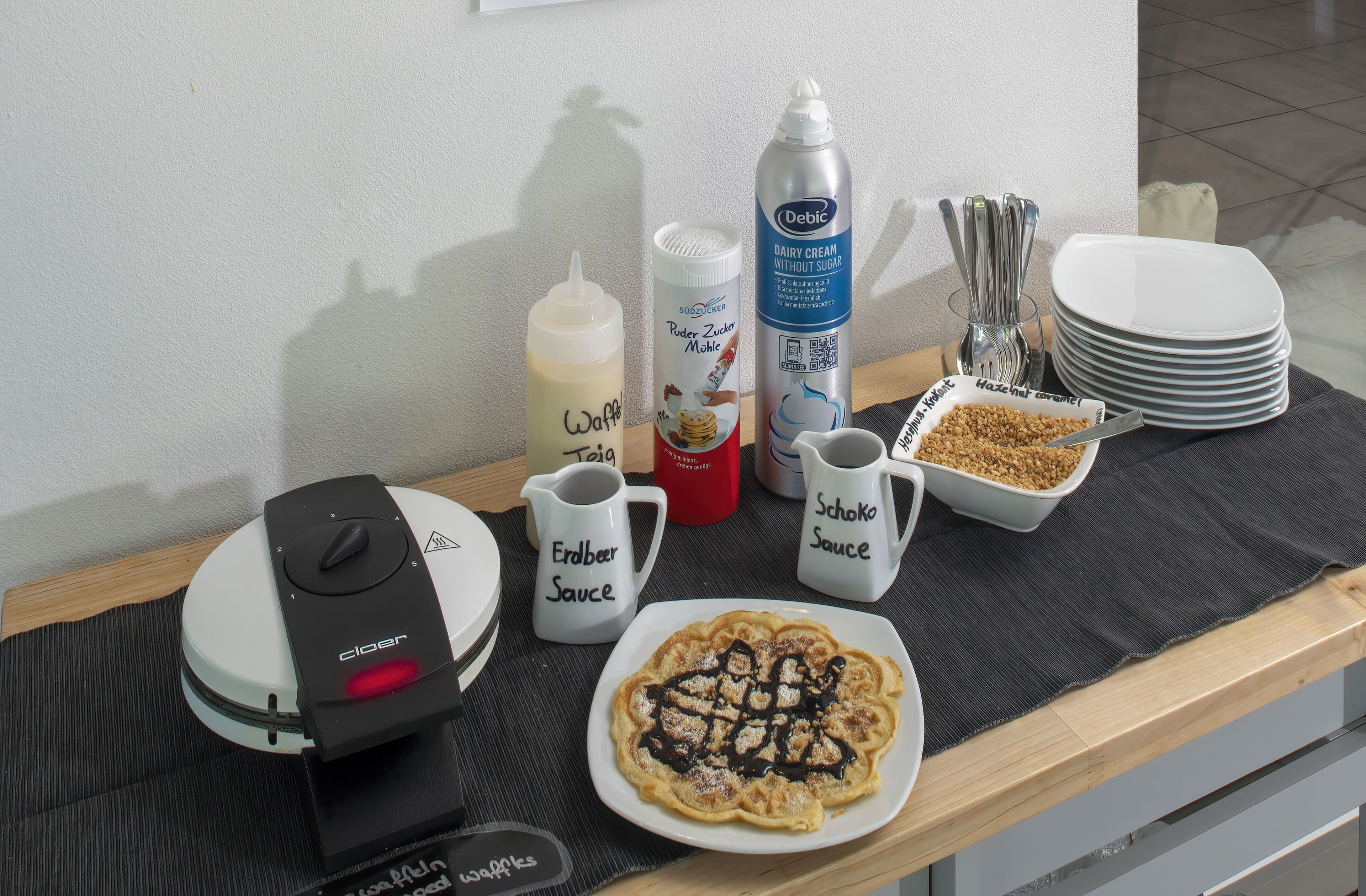 Uma mesa com café da manhã, incluindo waffles, molhos e uma máquina de waffles.