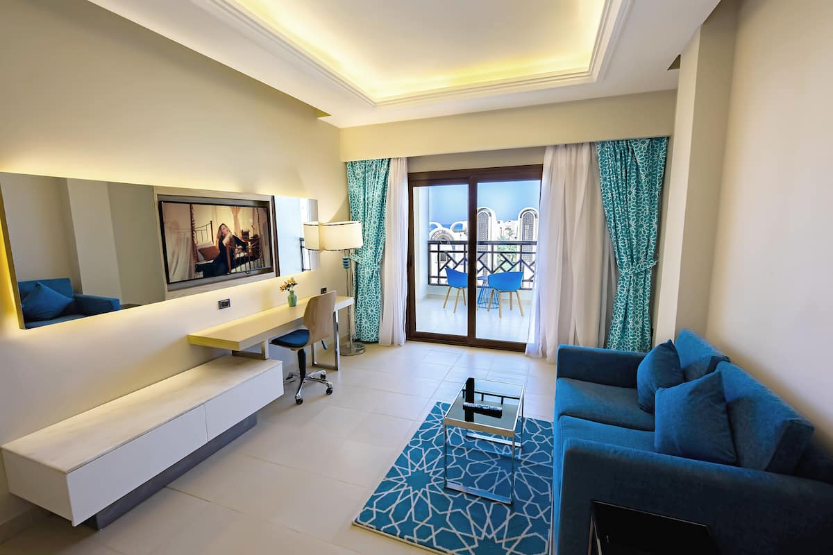 Quarto de hotel moderno com sofá, escrivaninha, cadeira, televisão na parede e varanda com área de refeições.
