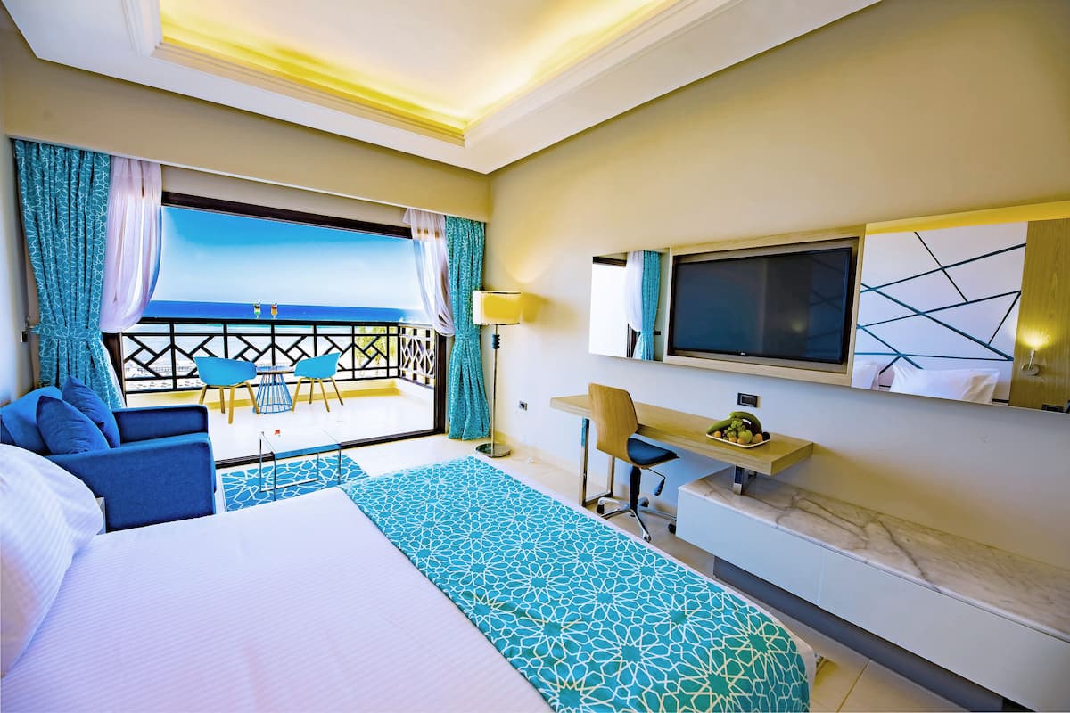 Quarto de hotel com cama, escrivaninha, cadeira, televisão e varanda com vista para o mar.
