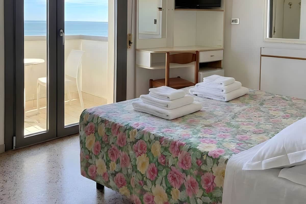 Quarto de hotel com cama, televisão, escrivaninha e varanda com cadeiras.
