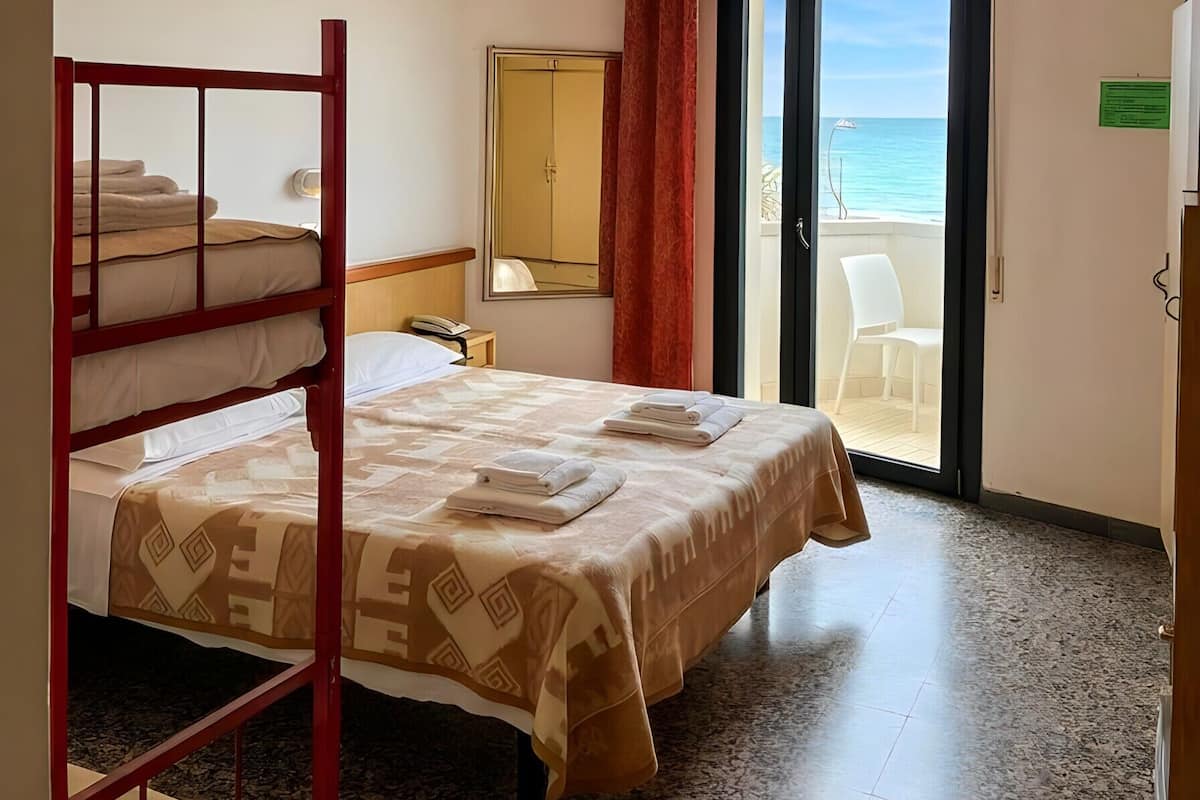 Quarto de hotel com beliche, cama de solteiro, grande janela com vista para o mar, porta que dá para uma varanda e luz no teto.