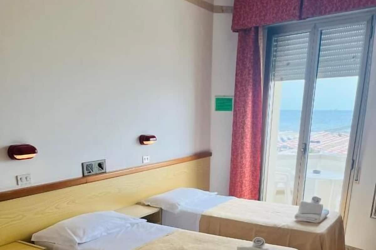 Quarto de hotel com três camas de solteiro, cada uma com uma toalha sobre a roupa de cama. Há uma janela com vista para a praia e uma cortina vermelha.