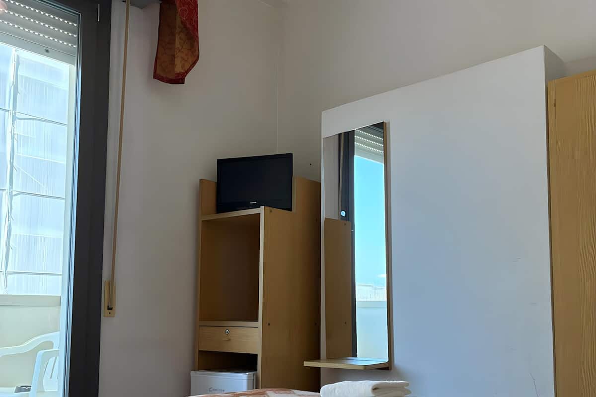 Quarto de hotel com uma cama, uma televisão sobre um suporte de madeira, um guarda-roupa e uma porta de correr para uma varanda.