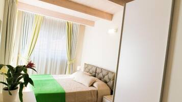 Quarto de hotel com cama, mesa de cabeceira, planta em vaso e janela com cortinas.