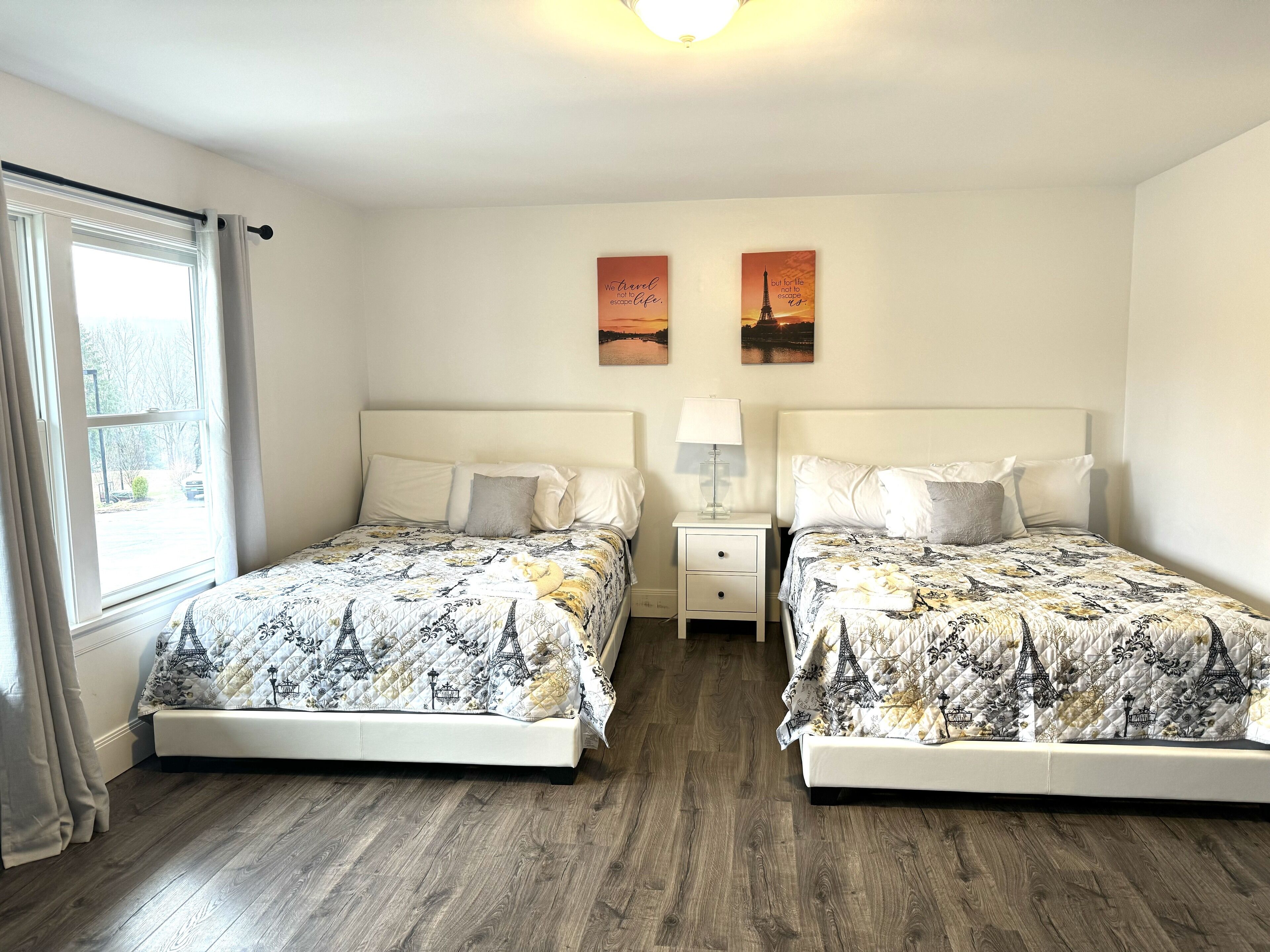 Quarto de casal com cama de casal, piso de madeira, duas mesas de cabeceira e uma janela com cortinas.