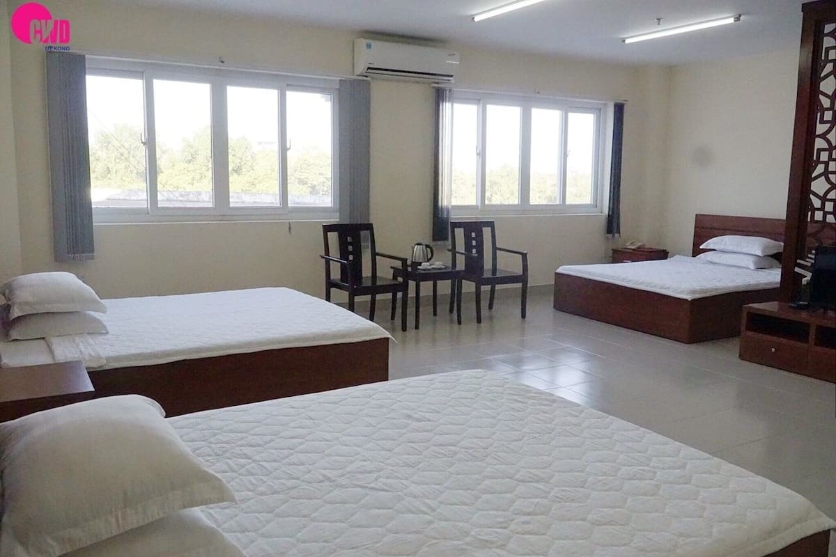 Quarto de hotel com duas camas de solteiro, uma pequena escrivaninha com cadeira e uma grande janela que oferece luz natural.