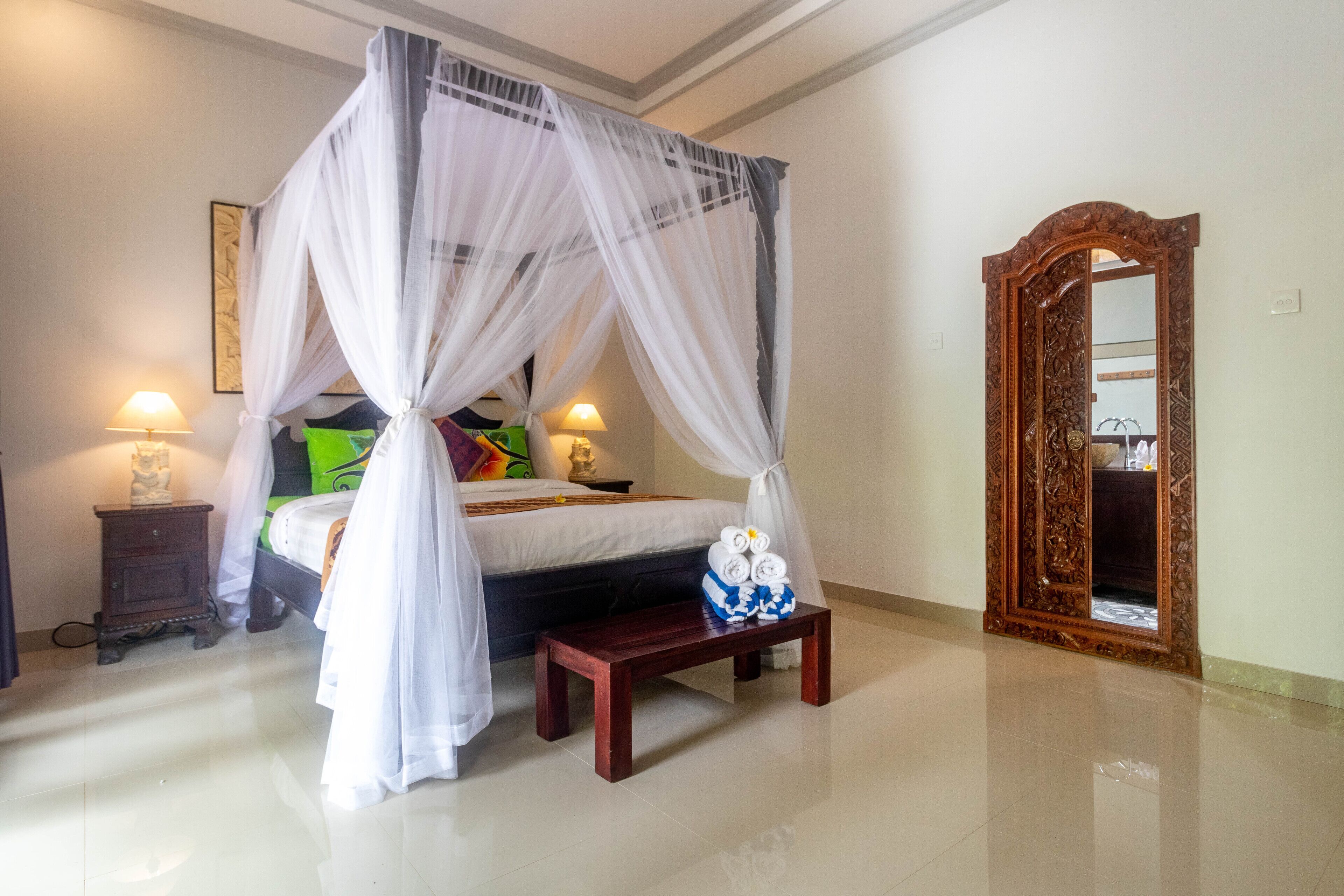Quarto com cama de dossel, um banco de madeira, um espelho e duas mesas de cabeceira com luminárias.