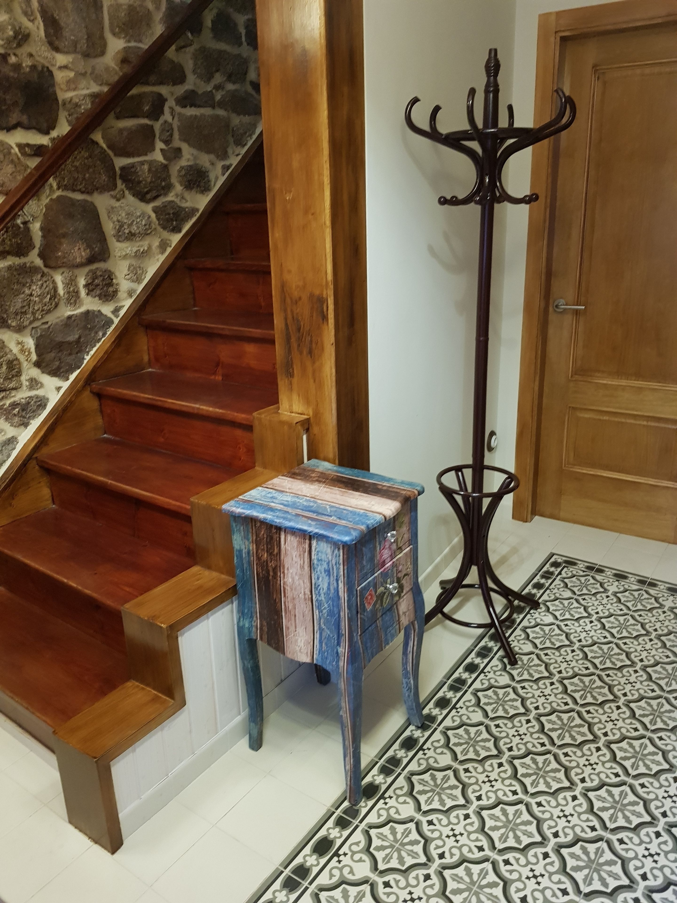 Uma escadaria com degraus de madeira e parede de pedra, uma pequena mesa de madeira com tampo de retalhos, um porta-roupas preto e um soalho estampado.