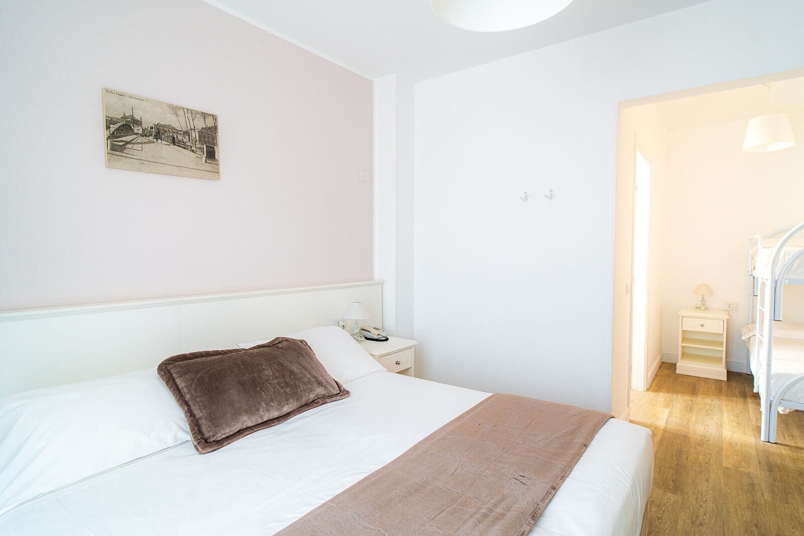 Um quarto com cama, mesa de cabeceira, abajur, um quadro na parede e uma beliche no quarto vizinho.