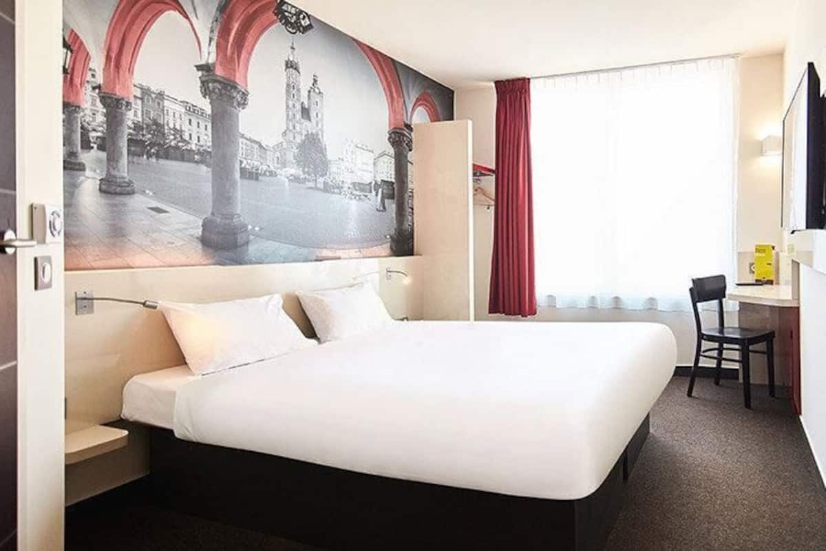 Quarto de hotel com uma cama grande, uma escrivaninha e uma cadeira. Há uma janela com cortinas, uma TV na parede e uma pintura de uma paisagem urbana.