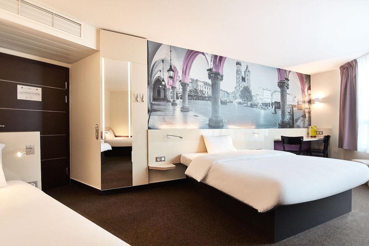 Quarto de hotel moderno com uma cama grande, área de estar e um mural com vista para a cidade na parede.