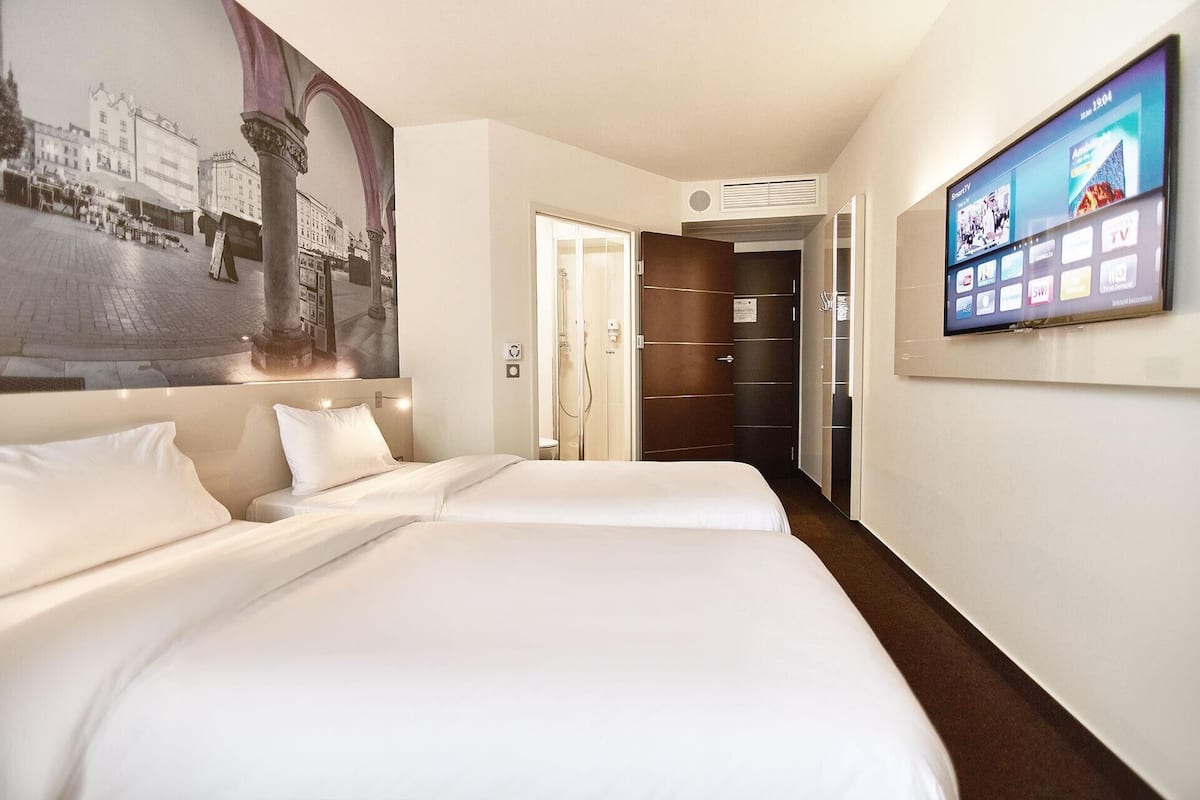 Quarto de hotel com duas camas, uma televisão grande na parede e um mural de uma paisagem urbana.