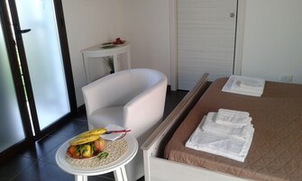 Quarto de hotel com uma cama, uma cadeira, uma mesinha com frutas e uma janela.