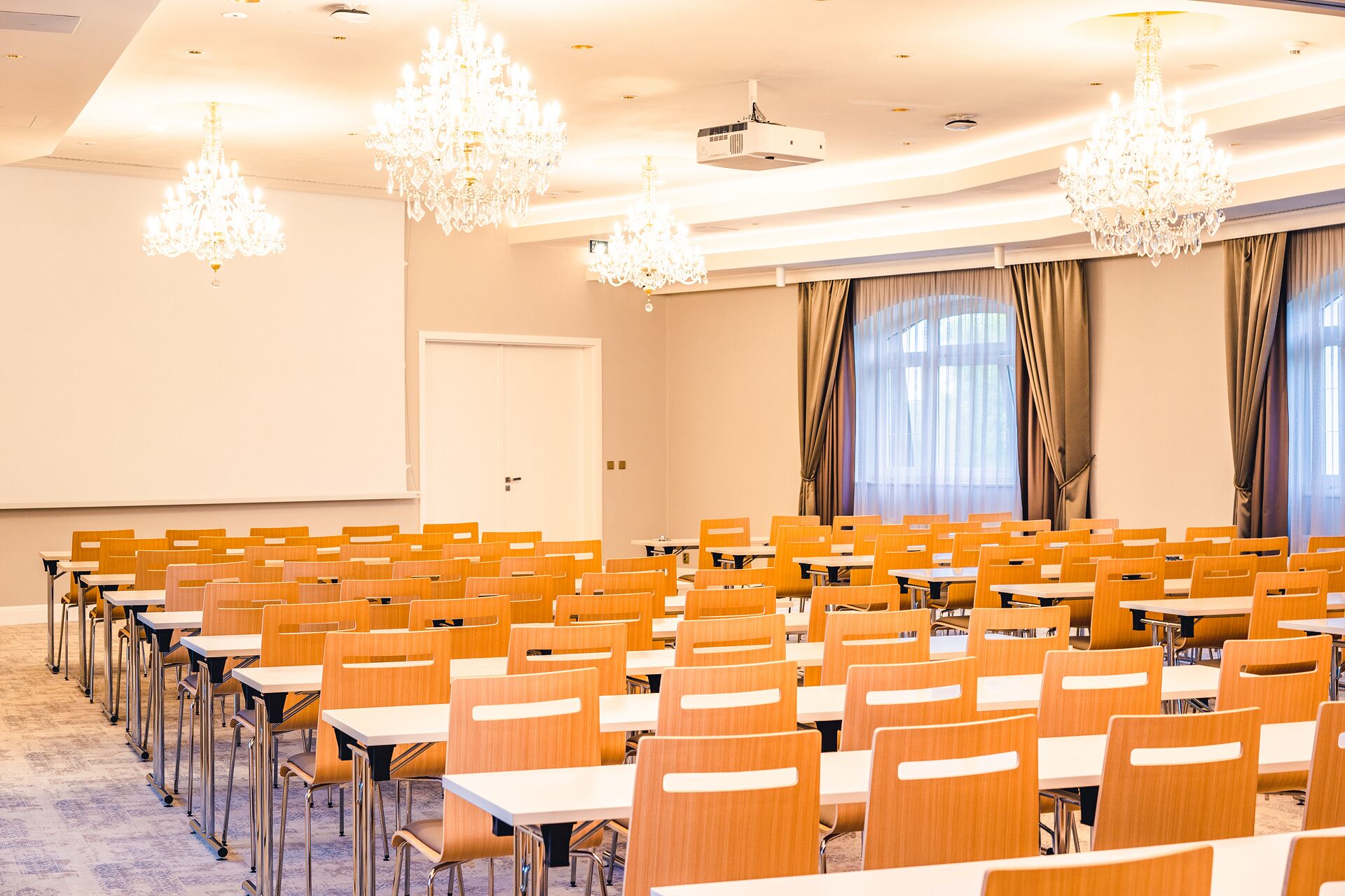 Uma sala de conferências com fileiras de cadeiras e mesas de madeira, janelas amplas com cortinas e lustres.
