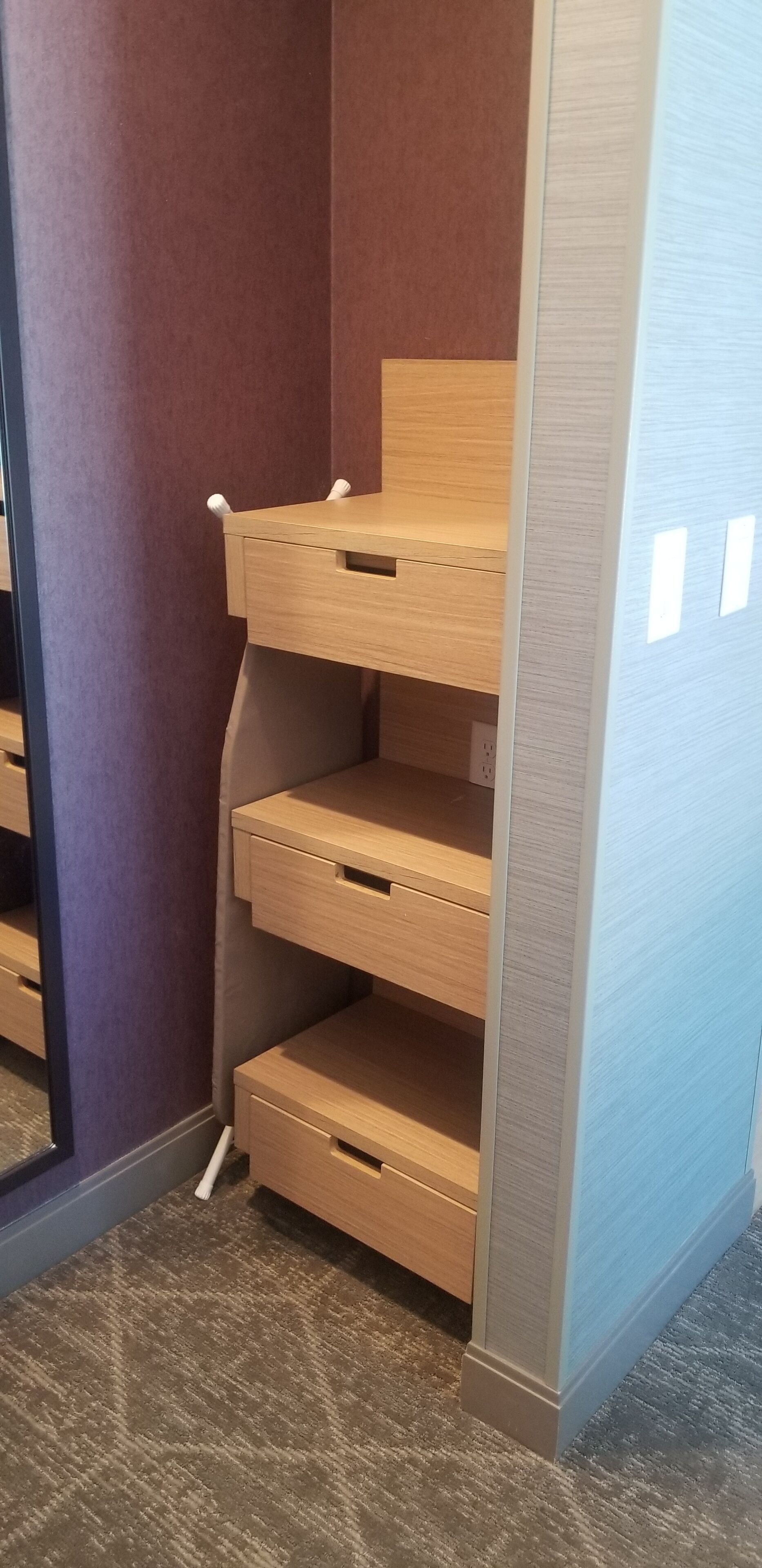 Canto de um quarto com uma estante de madeira encostada a uma parede com painel roxo e azul claro.