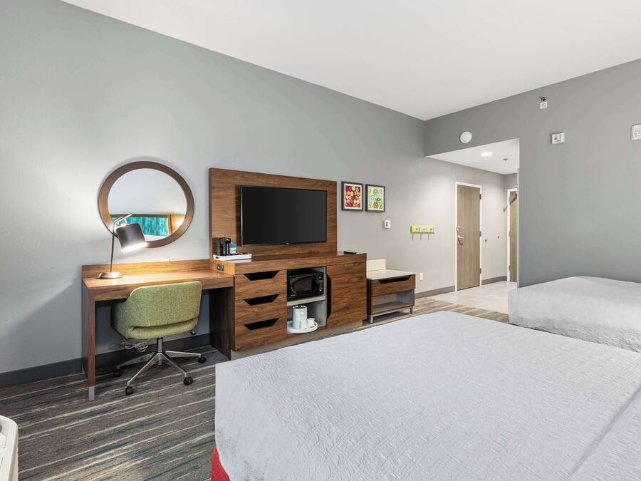Quarto de hotel com escrivaninha de madeira, televisão de tela plana, cadeira de escritório verde, micro-ondas, pequeno armário e duas camas de solteiro.