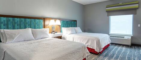 Quarto de hotel com duas camas, uma mesinha de cabeceira com abajur, uma janela com ar condicionado e vista para o exterior.