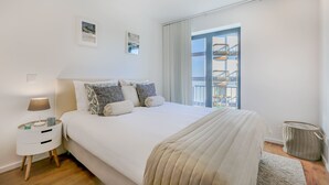 Quarto moderno com cama grande, mesa de cabeceira com abajur e vista para um edifício pela janela.