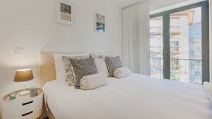 Quarto de hotel com uma cama grande, uma mesinha de cabeceira com abajur e duas fotografias emolduradas na parede.