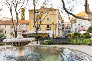 Uma fonte em praça pública, com bancos e um edifício ao fundo.