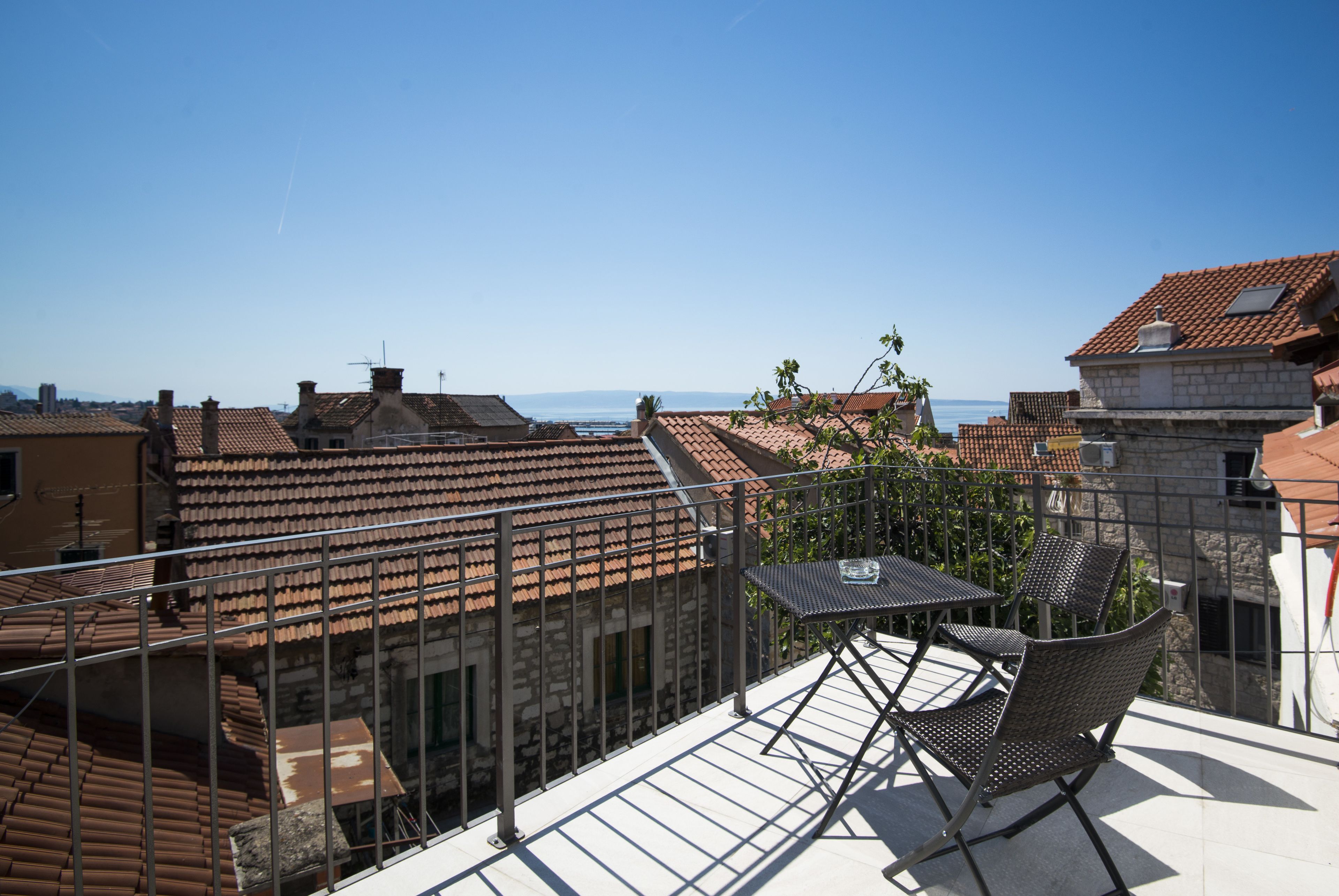 Terraço no telhado com mesa e cadeiras, vista para uma paisagem urbana com edifícios de telhado em terracota e céu azul límpido.