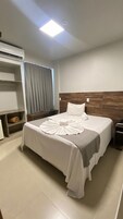 Cama bem arrumada com colcha branca e almofadas, cabeceira de madeira, mesa de cabeceira e prateleira com toalha enrolada.