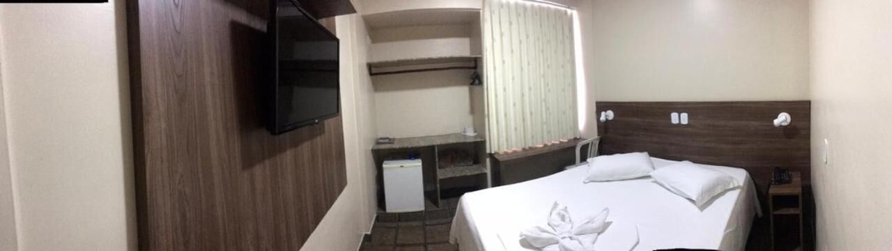 Quarto de hotel com cama, uma pequena escrivaninha, televisão e janela com cortinas.