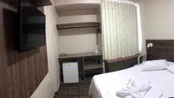 Quarto de hotel com cama, uma pequena escrivaninha, televisão e janela com cortinas.