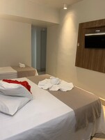 Quarto de hotel com duas camas, cada uma com lençóis brancos e um cobertor vermelho em uma delas. Há uma televisão fixada na parede e uma porta que dá para outro quarto.
