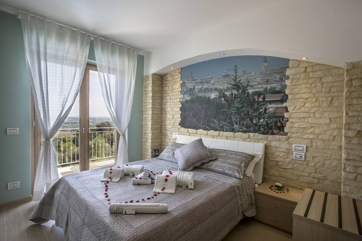 Quarto com uma cama grande, vista para a cidade através de uma janela e uma parede de pedra.